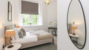 Quarto com cama, abajures, escrivaninha, cadeira, espelho grande e janela com vista para árvores.