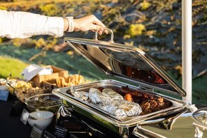 Uma mesa de buffet com diversos tipos de comida, incluindo itens embalados em papel alumínio e uma churrasqueira com alimentos sendo preparados.