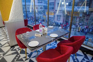 Restaurante com mesa posta para dois, cadeiras vermelhas e vista para a rua através de grandes janelas.