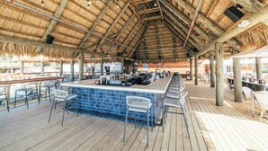 Uma área de bar ao ar livre coberta, com deck de madeira, telhado de palha e banquetas.