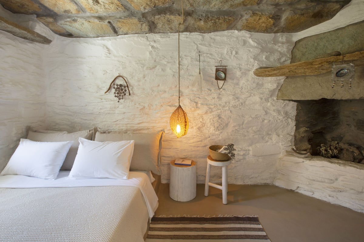 Um quarto aconchegante com parede de pedra, cama de madeira, luminária pendente e um pequeno banquinho de madeira.