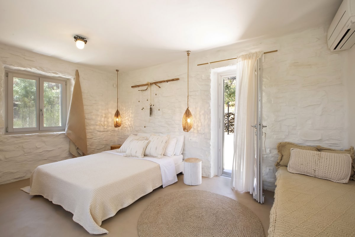 Quarto com uma cama grande, uma mesinha de cabeceira, um tapete redondo e uma janela com cortinas.