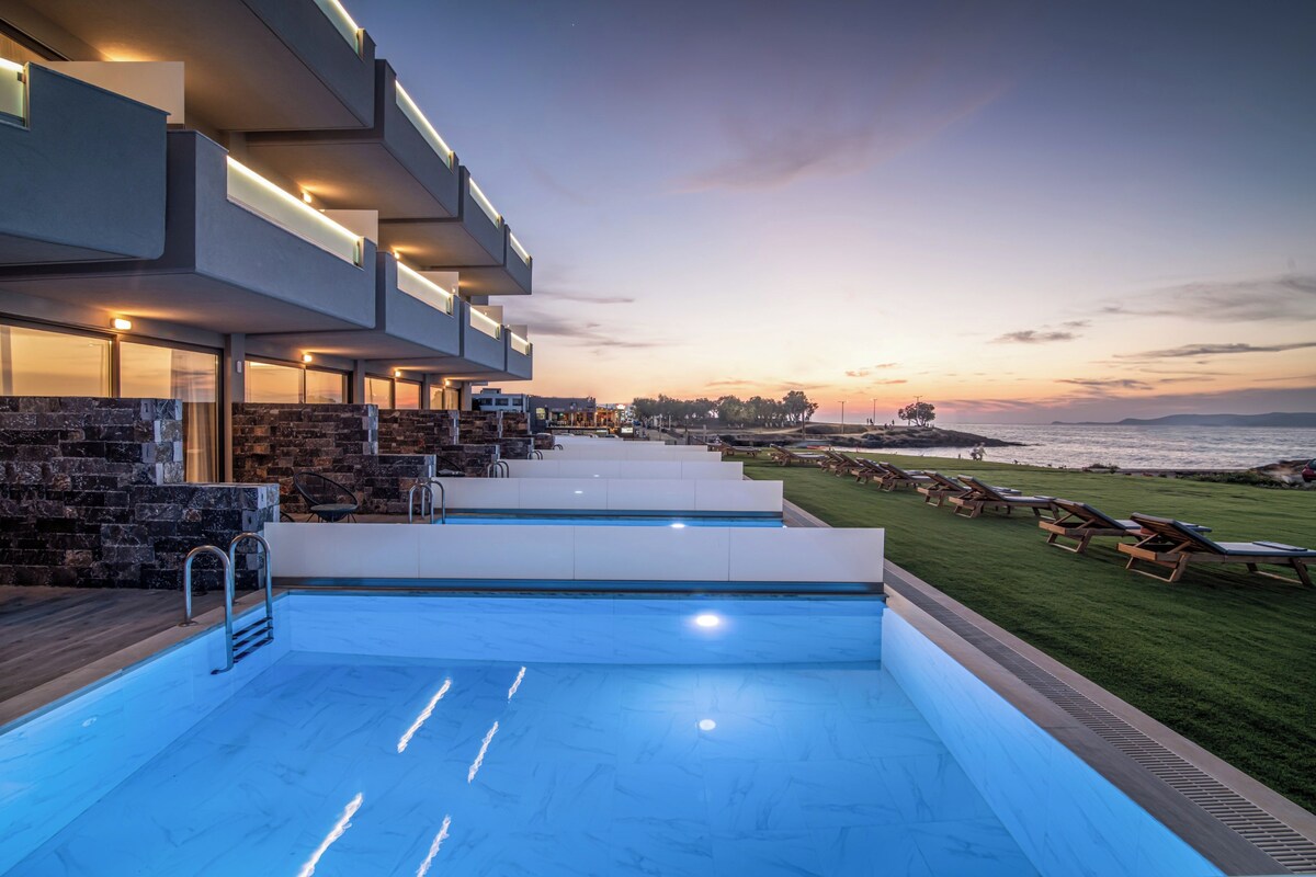 Uma piscina com vista para o oceano ao pôr do sol, um edifício moderno com varandas e espreguiçadeiras na relva.