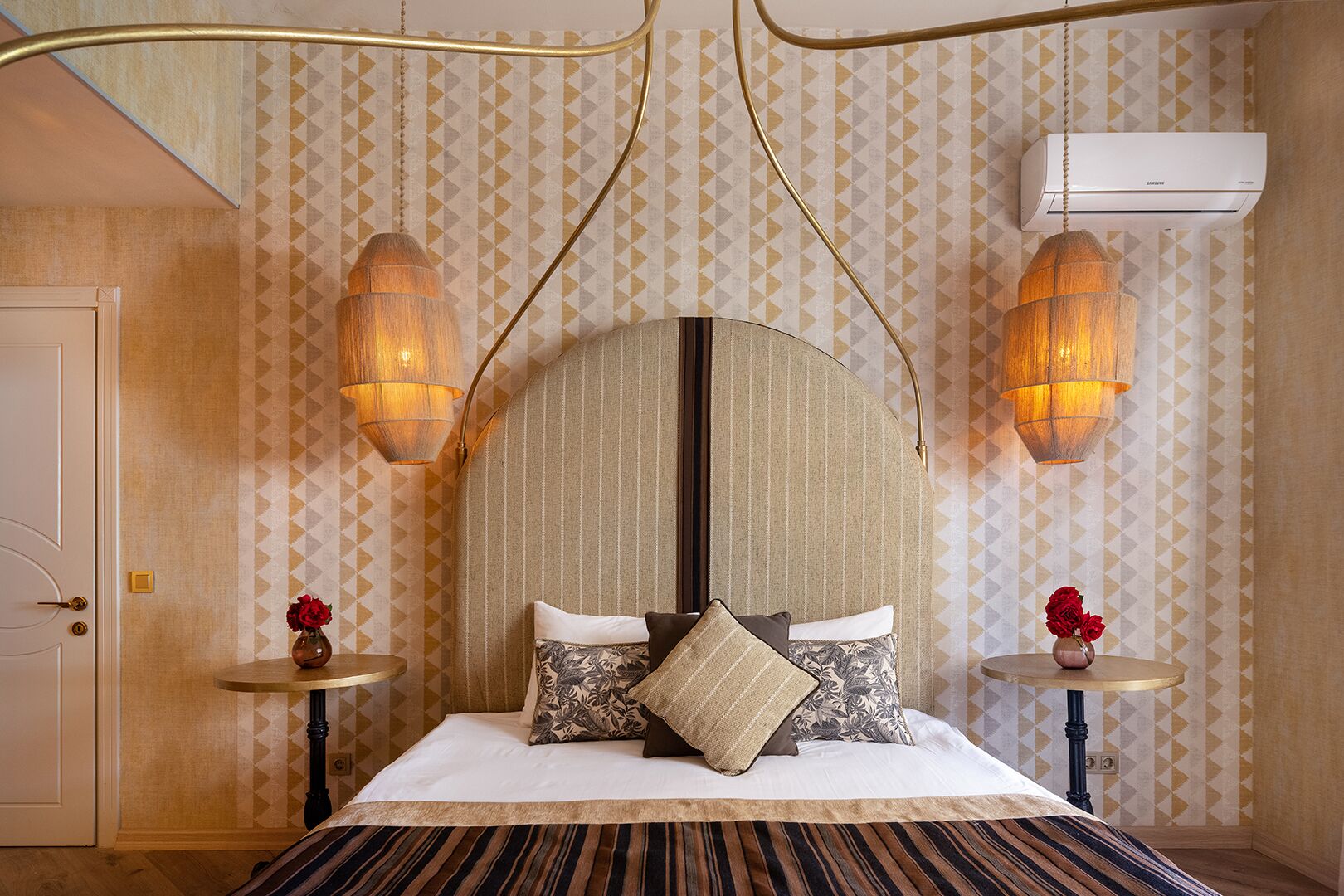 Quarto de hotel com uma cama grande, duas mesas de cabeceira com abajures, ar-condicionado fixado na parede e papel de parede estampado.
