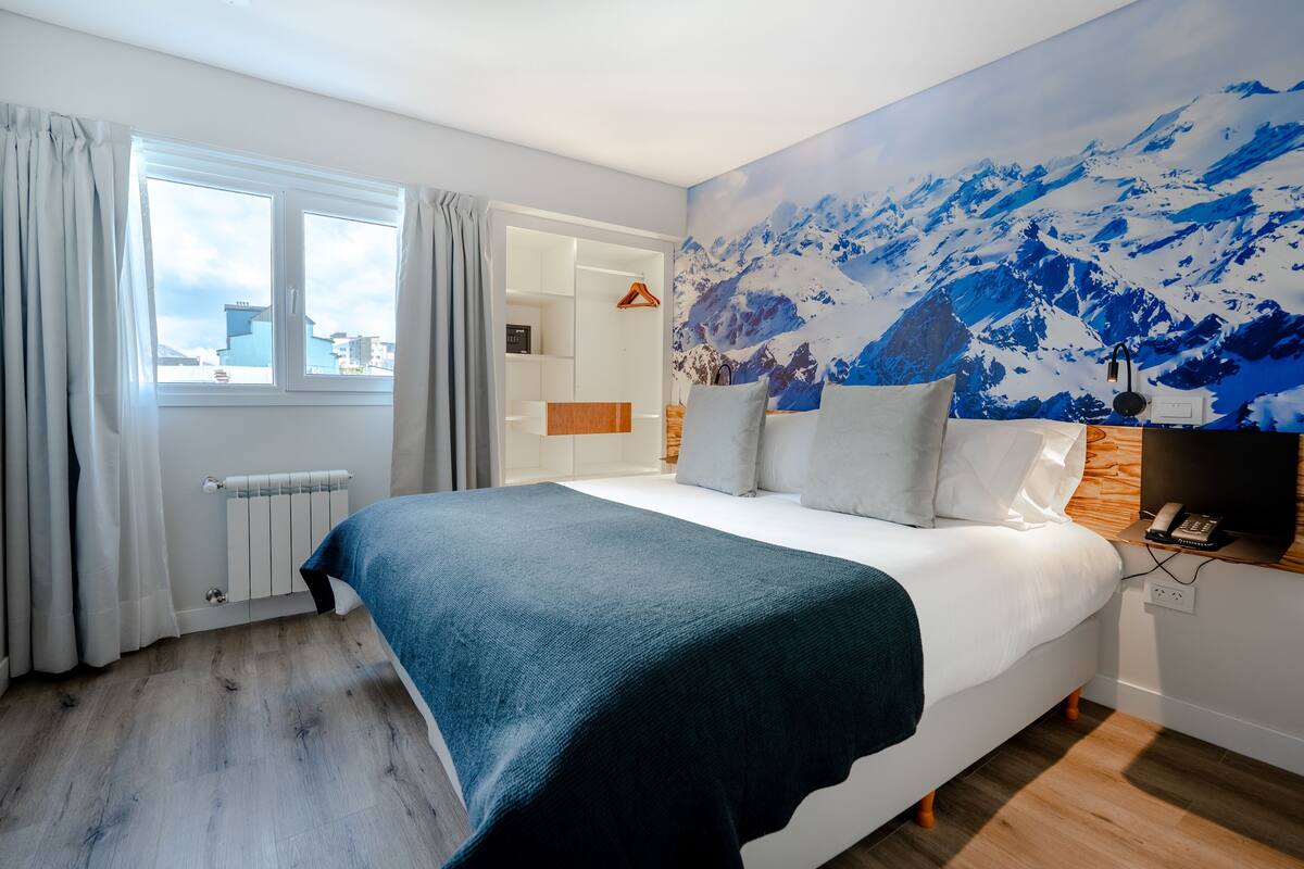 Quarto com cama, janela, radiador, TV fixada na parede e um mural de montanha.