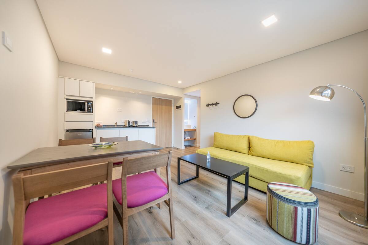 Uma sala de estar moderna com um sofá amarelo, uma mesa de jantar de madeira com almofadas rosa, uma mesa de centro preta e um banquinho listrado.