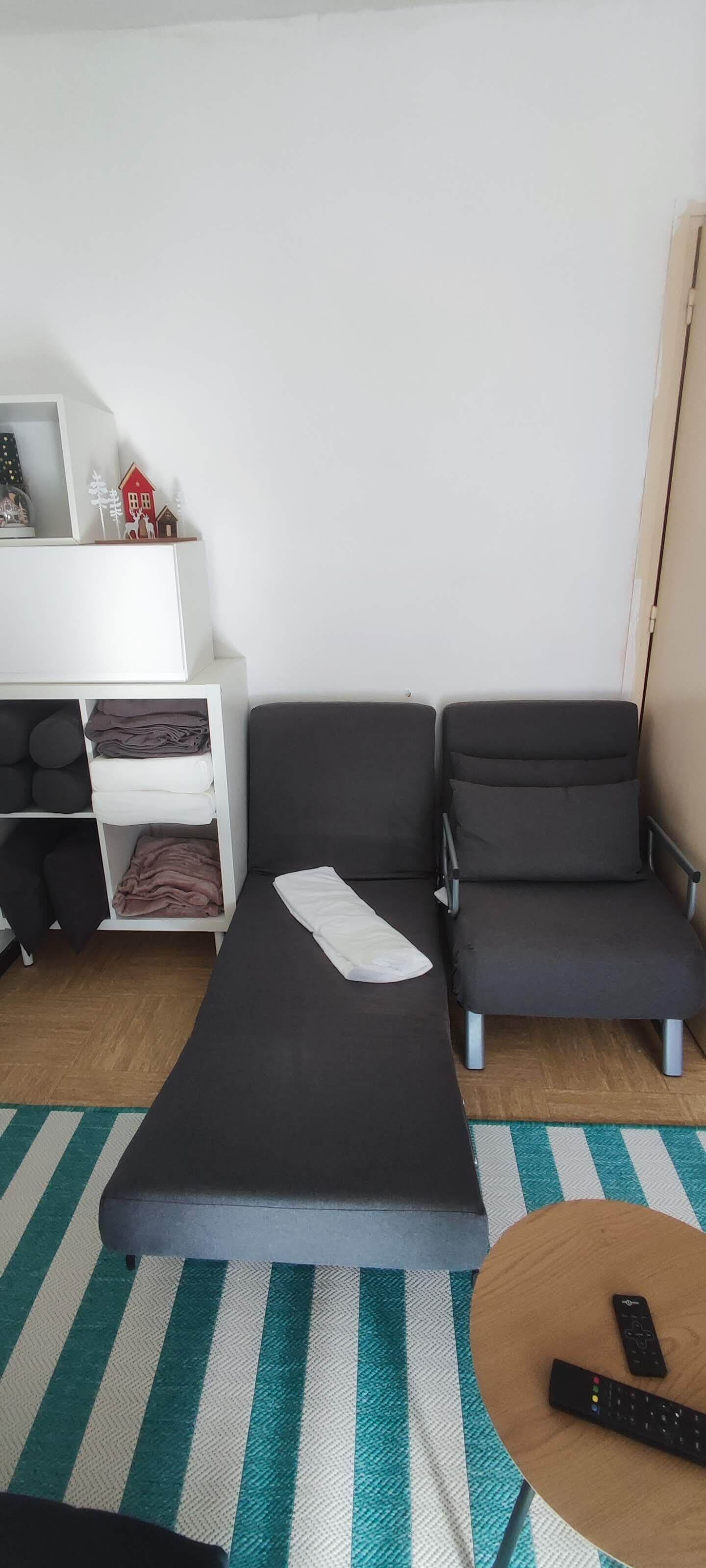 Quarto com sofá, uma pequena mesa com um controle remoto e uma estante branca com objetos dobrados e uma miniatura decorativa de casa.