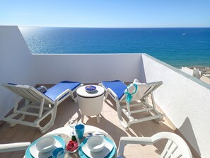 Terraço com mobiliário branco, mesa posta com pratos, tigelas e um copo de água, com vista para o mar.