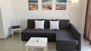 Uma sala de estar moderna com um sofá modular cinza, duas almofadas brancas, uma mesinha branca e uma vela de vidro. Há três quadros na parede retratando cenas de praia. Uma cadeira branca e um abajur também estão visíveis.