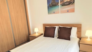Quarto com uma cama, duas mesas de cabeceira com abajures, um guarda-roupa de madeira e um quadro na parede retratando uma praia.