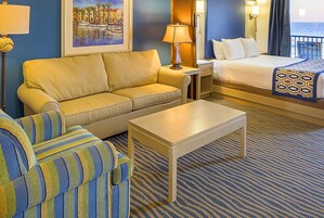 Quarto de hotel com sofá, poltrona listrada, cama, mesa de cabeceira, luminária e pintura de uma paisagem costeira.