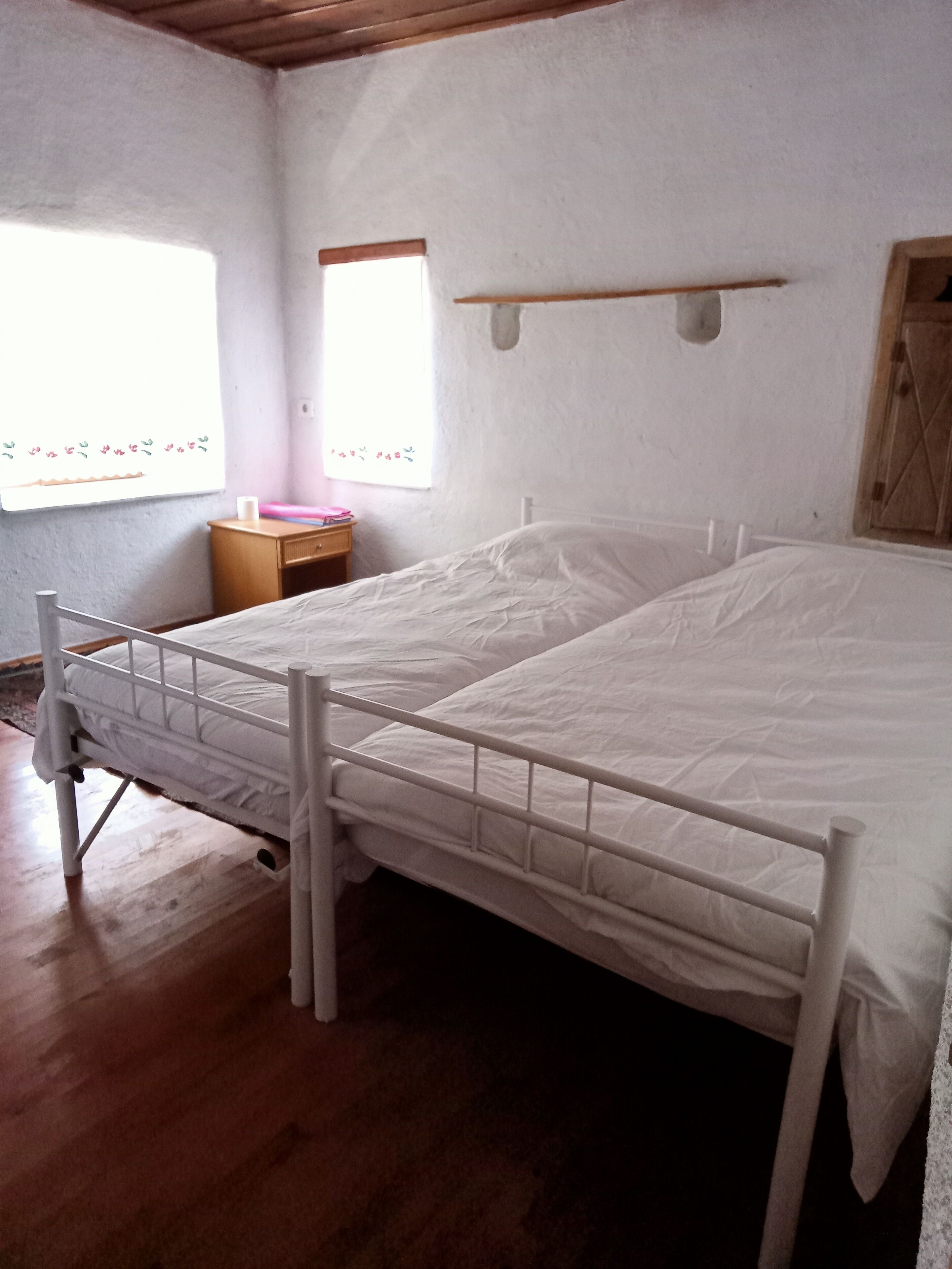 Quarto individual com uma cama, piso de madeira, paredes brancas, guarda-roupa de madeira, mesa de cabeceira pequena e janela com vista para o exterior.