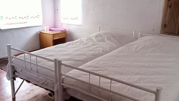 Quarto individual com uma cama, piso de madeira, paredes brancas, guarda-roupa de madeira, mesa de cabeceira pequena e janela com vista para o exterior.