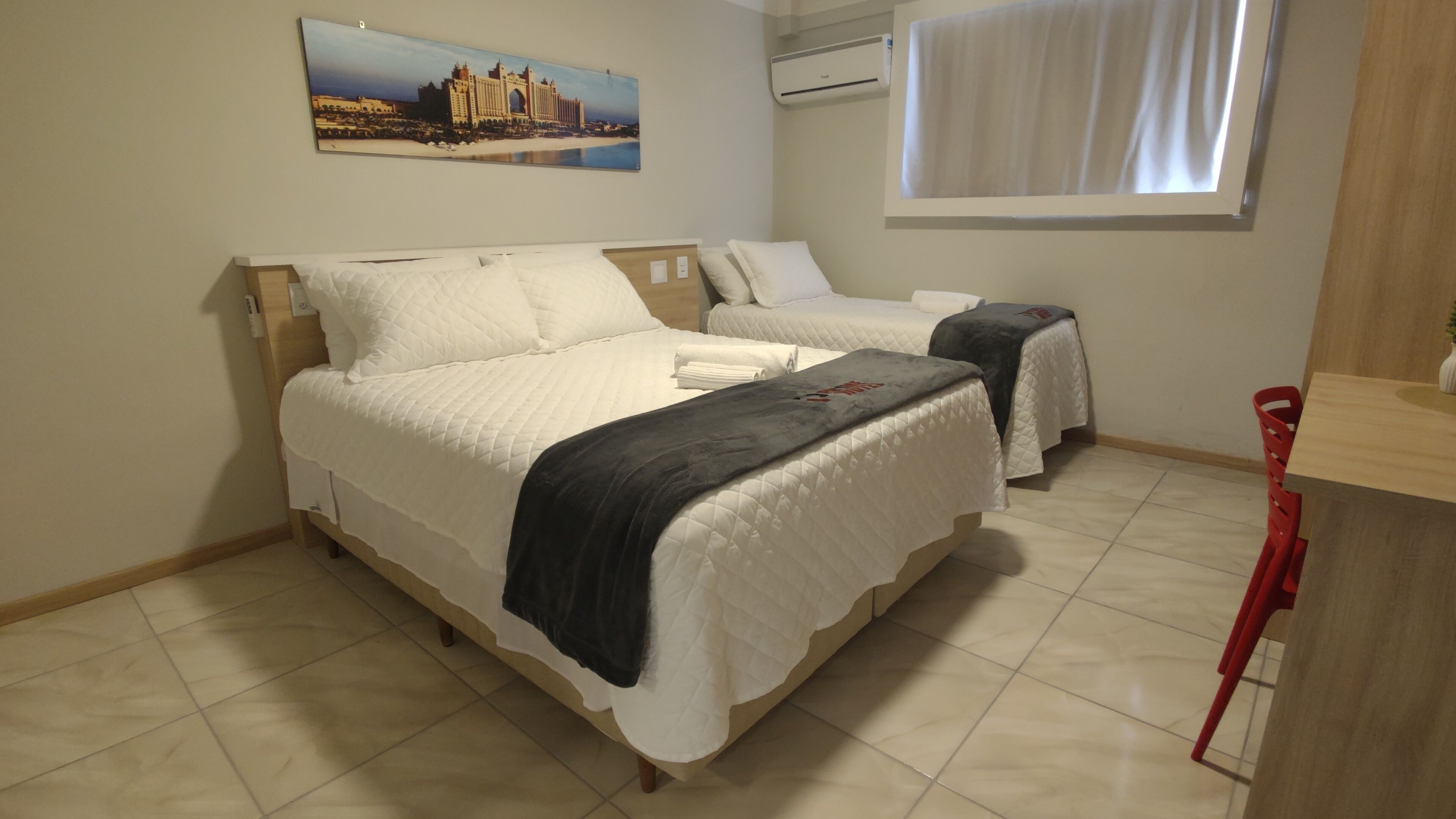 Quarto de hotel com duas camas, um quadro de um ponto turístico, um aparelho de ar condicionado e uma janela com cortinas.