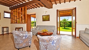 Uma sala de estar com sofá, duas poltronas, mesa de centro, vigas de madeira no teto, um quadro e vista através de uma porta de vidro.