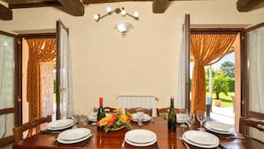 Mesa de jantar posta com pratos, copos e garrafas, rodeada por cadeiras, com vista para o verde lá fora.