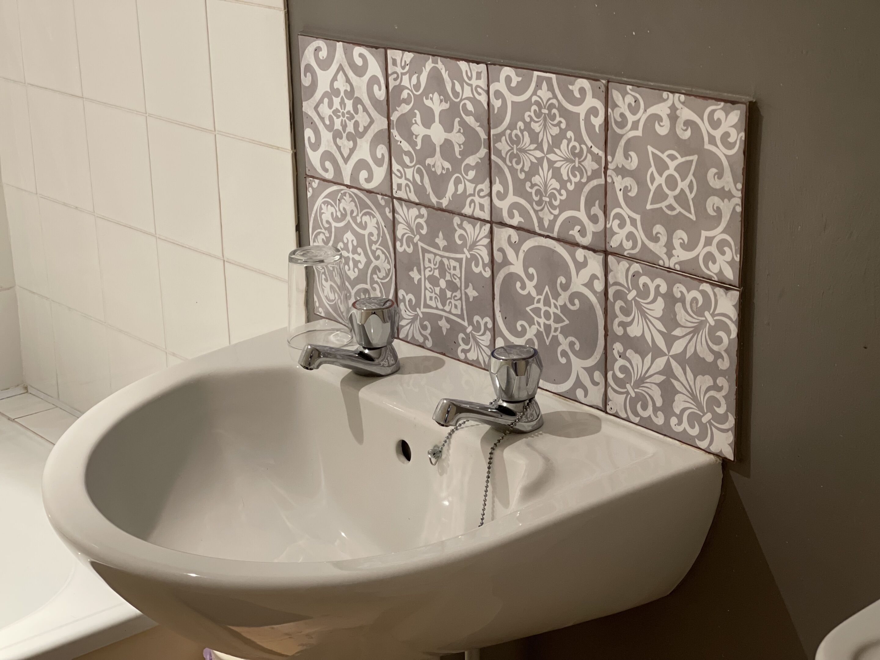 Casa de banho com lavatório branco, duas torneiras e painel decorativo de azulejos.