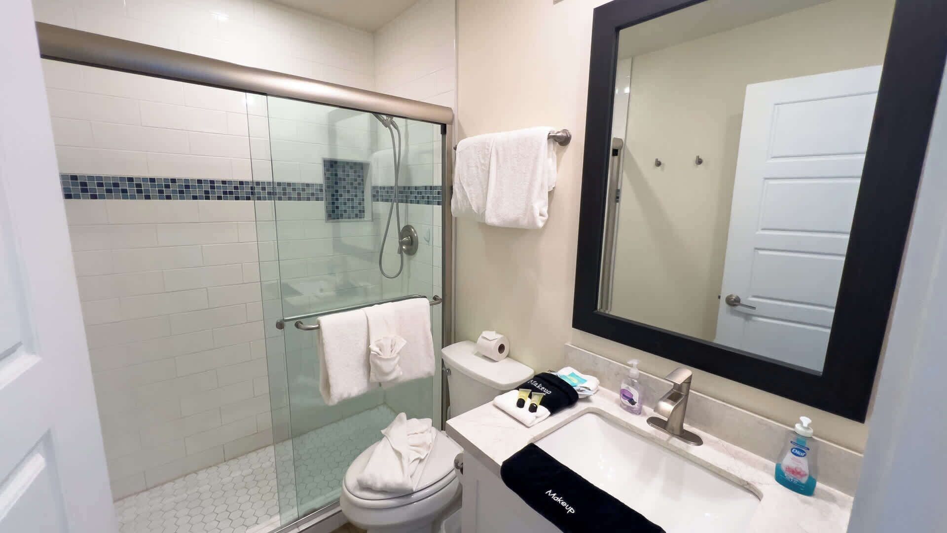 Uma casa de banho moderna com cabine de duche em vidro, sanita, lavatório com espelho para maquilhar e porta-toalhas.