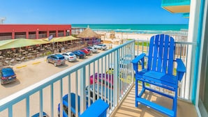 Uma varanda com uma cadeira azul, vista para a praia, um estacionamento com carros e um edifício com fachada vermelha.