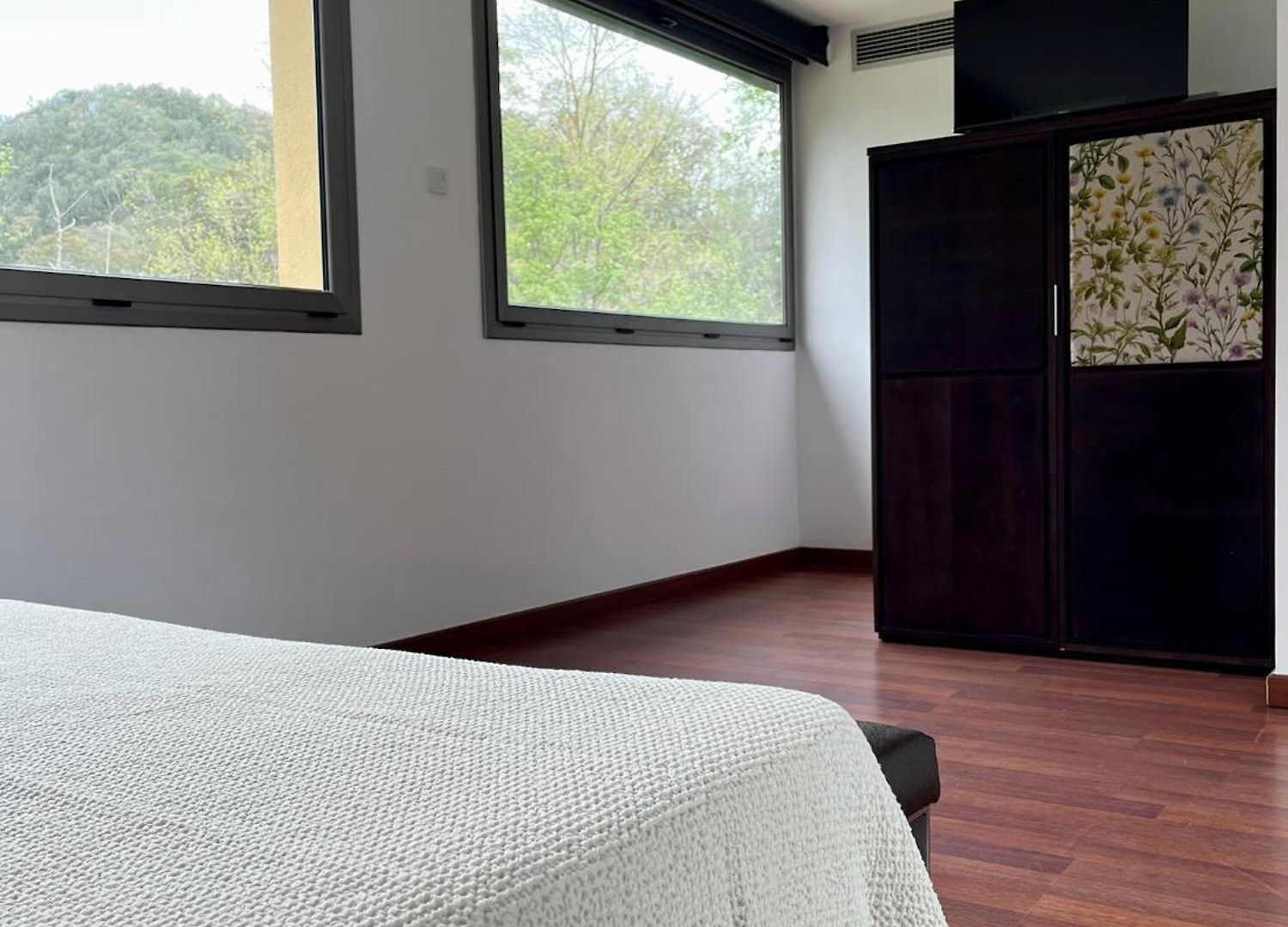 Quarto com cama, duas janelas, guarda-roupa e vista para árvores.