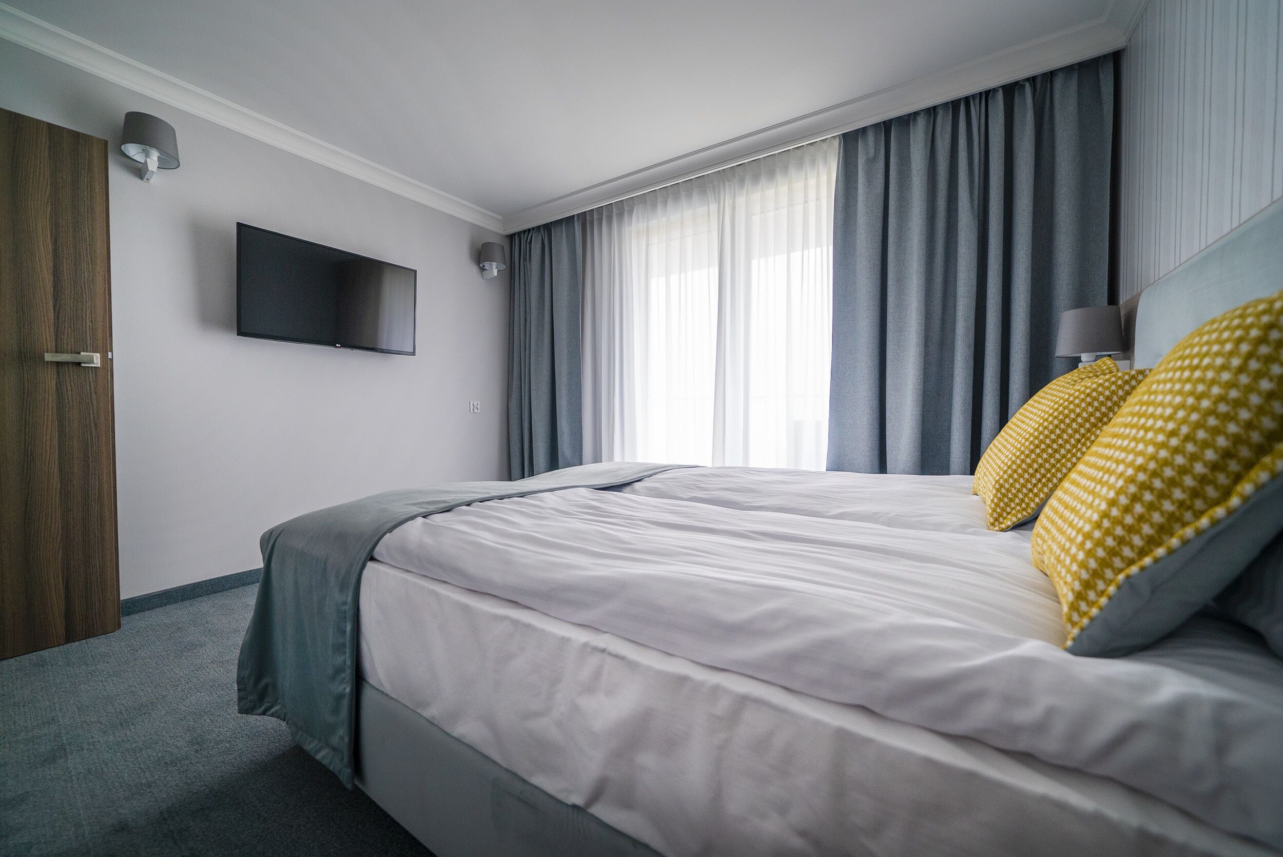 Quarto de hotel moderno com uma cama grande, televisão de tela plana na parede e uma janela com cortinas.