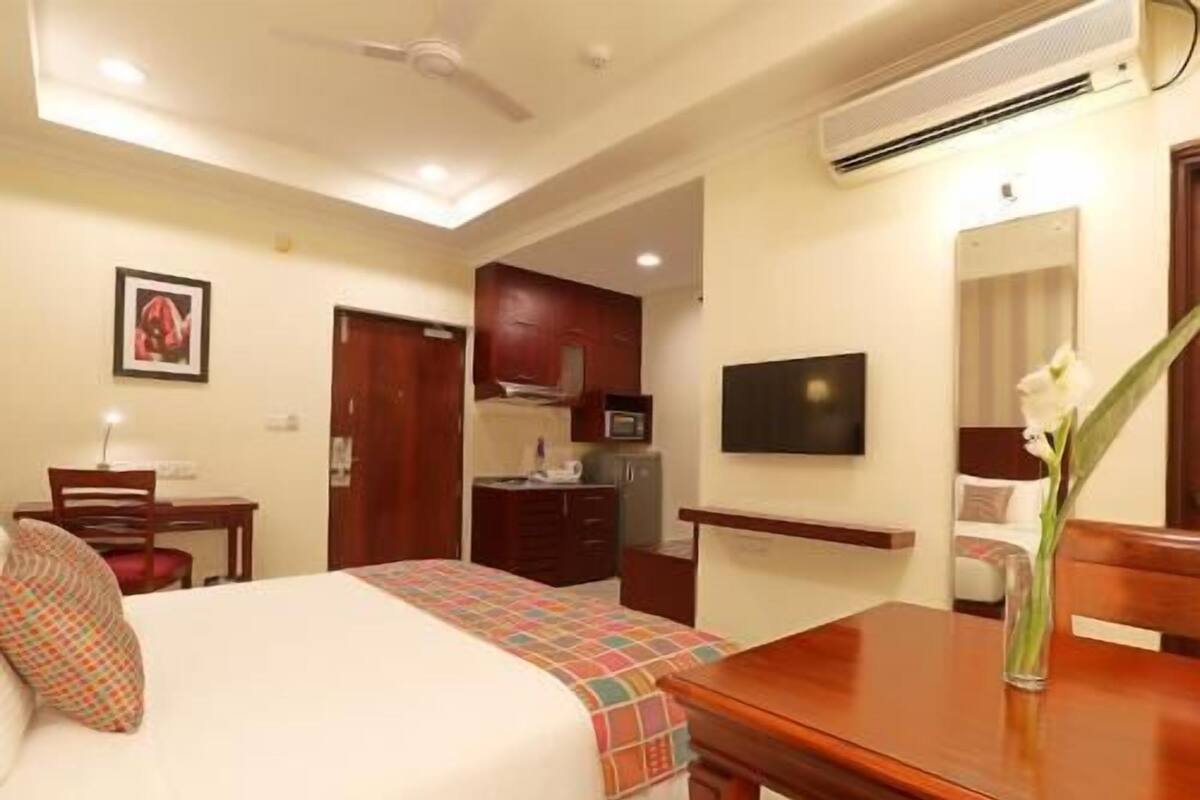 Quarto de hotel com cama, escrivaninha, cadeira, televisão, espelho e uma pequena cozinha compacta.