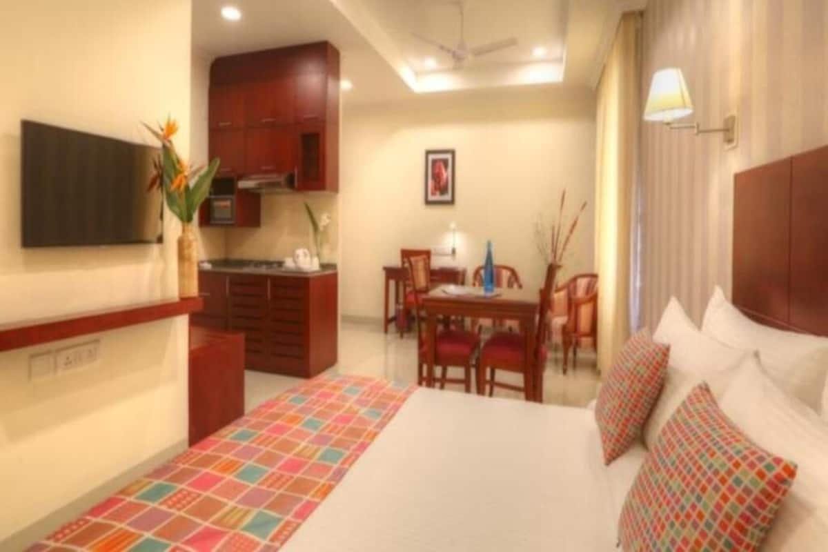 Quarto de hotel com cama, escrivaninha, cadeira, televisão e cozinha compacta.