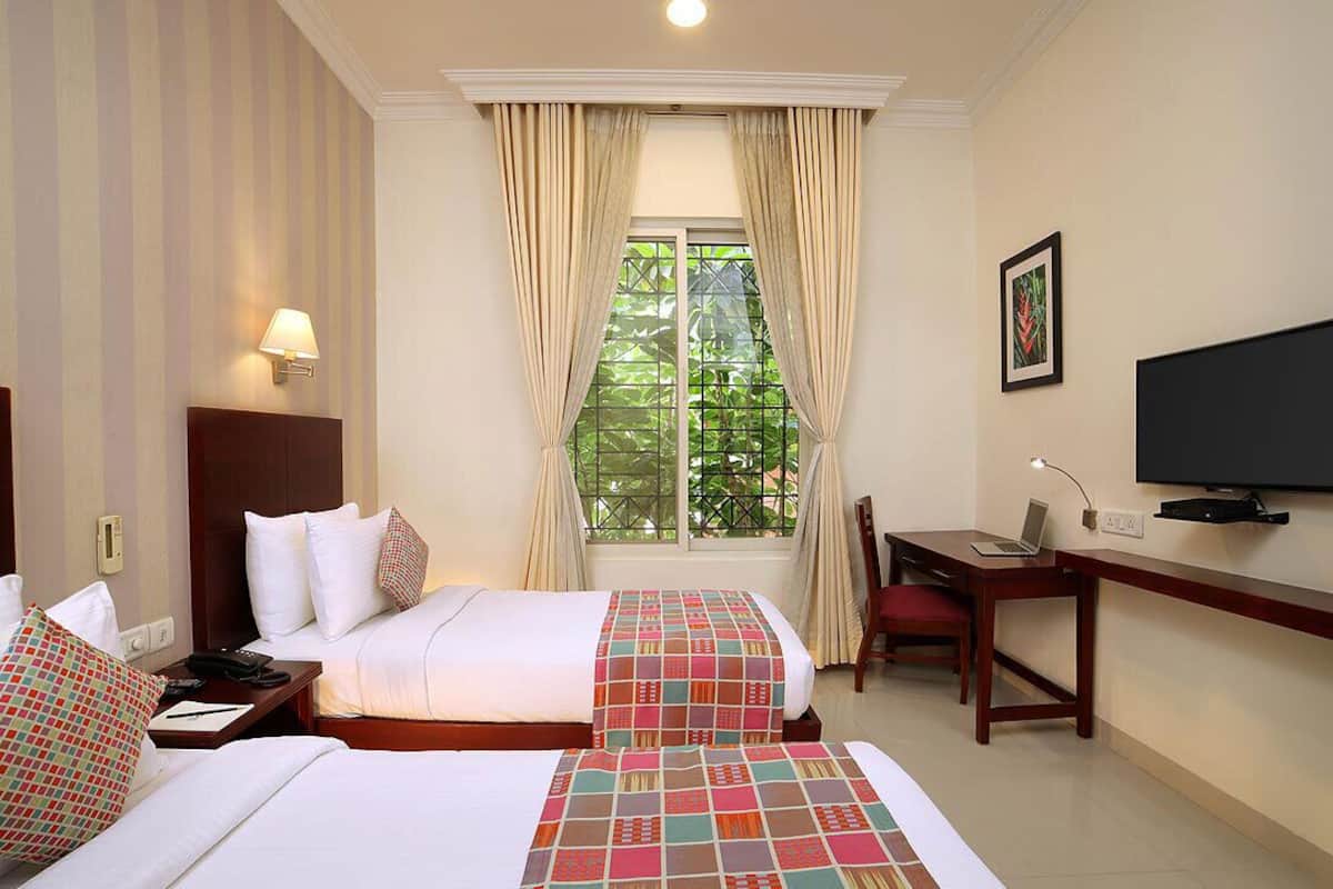 Quarto de hotel com duas camas, uma escrivaninha, uma cadeira, televisão e janela com cortinas.