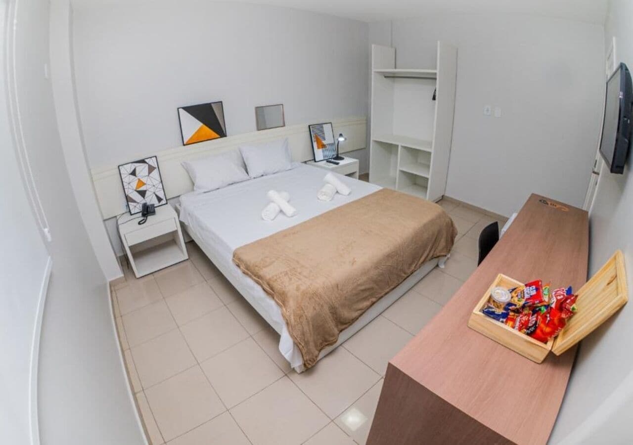Quarto de hotel com cama, mesas de cabeceira, escrivaninha de madeira e televisão.