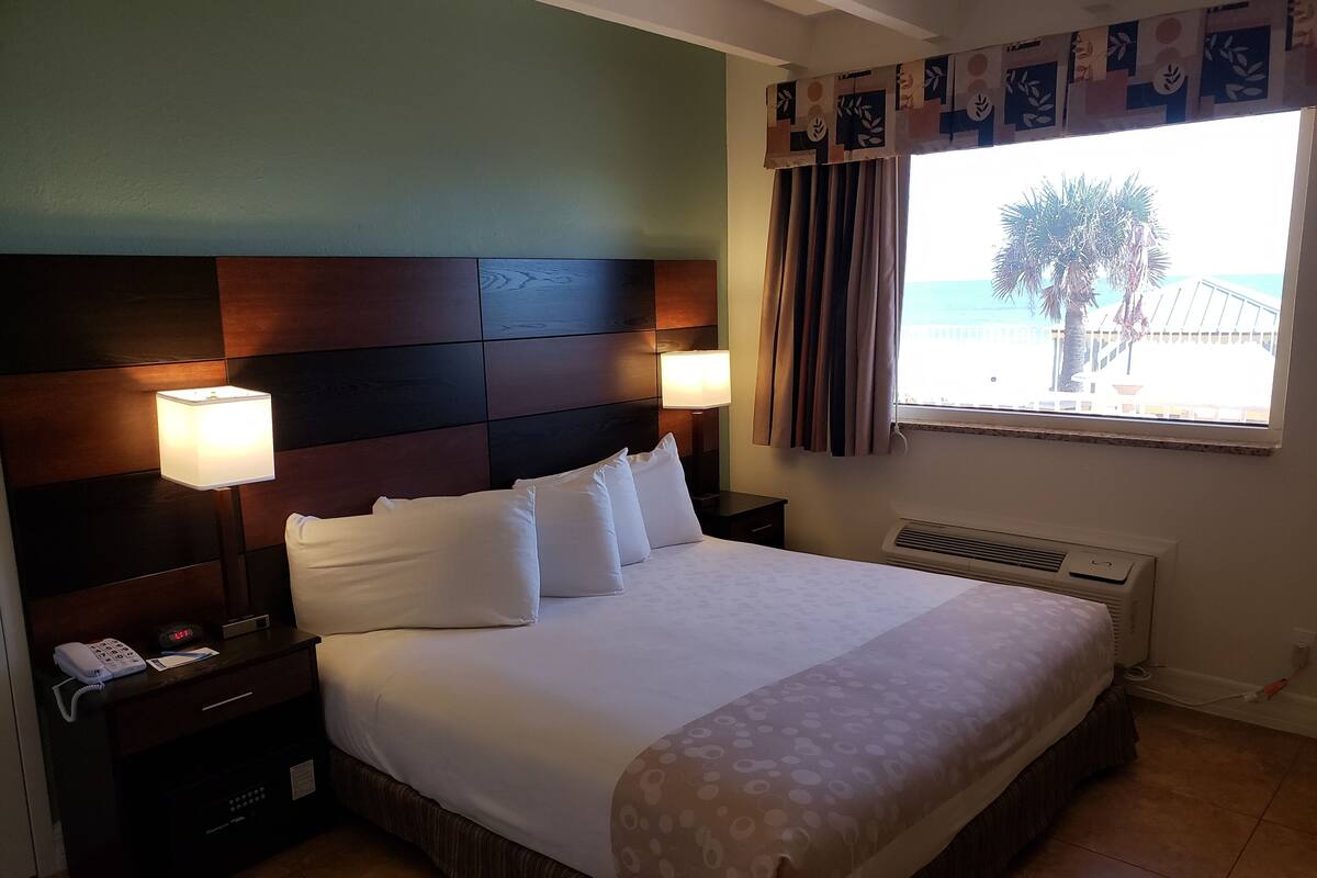 Quarto de hotel com uma cama grande, duas luminárias de cabeceira, um telefone e uma janela com vista para palmeiras e o mar.