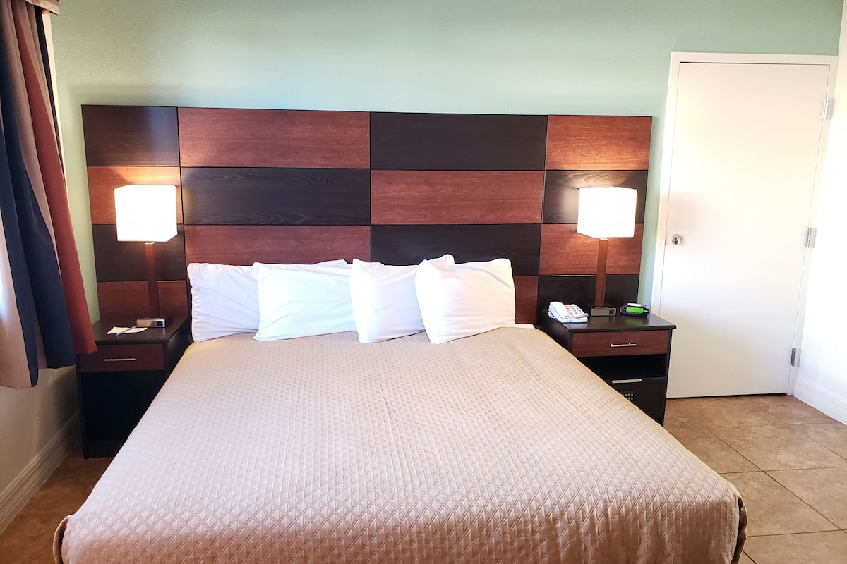 Quarto de hotel com uma cama grande, duas mesas de cabeceira, um criado-mudo, um abajur e uma porta.