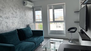 Uma sala de estar moderna com um sofá verde-turquesa, uma mesa de centro de vidro, uma TV de tela plana e uma grande janela com persianas.