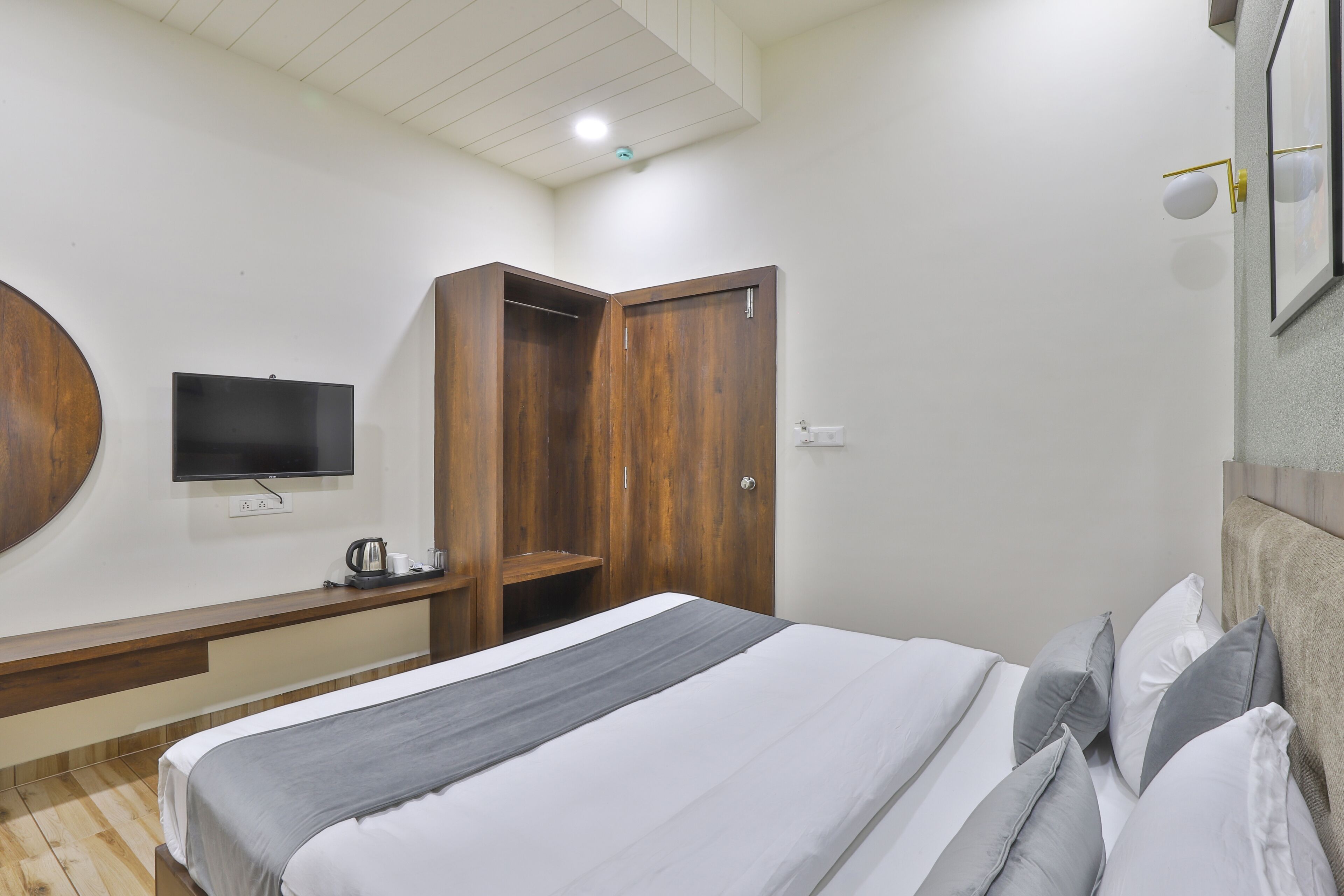 Quarto com cama, televisão na parede, guarda-roupa de madeira e mesa de cabeceira com chaleira.
