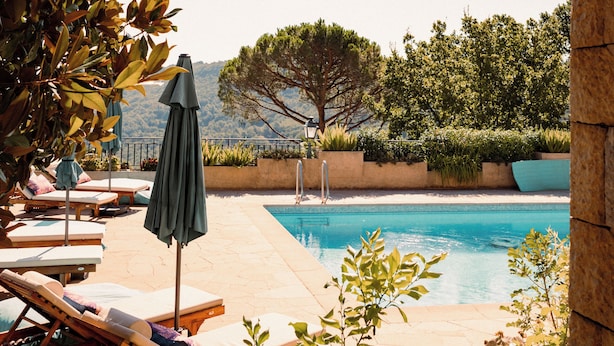 Uma área à beira da piscina com espreguiçadeiras e guarda-sóis, com vista direta para a piscina, e um arco de pedra.