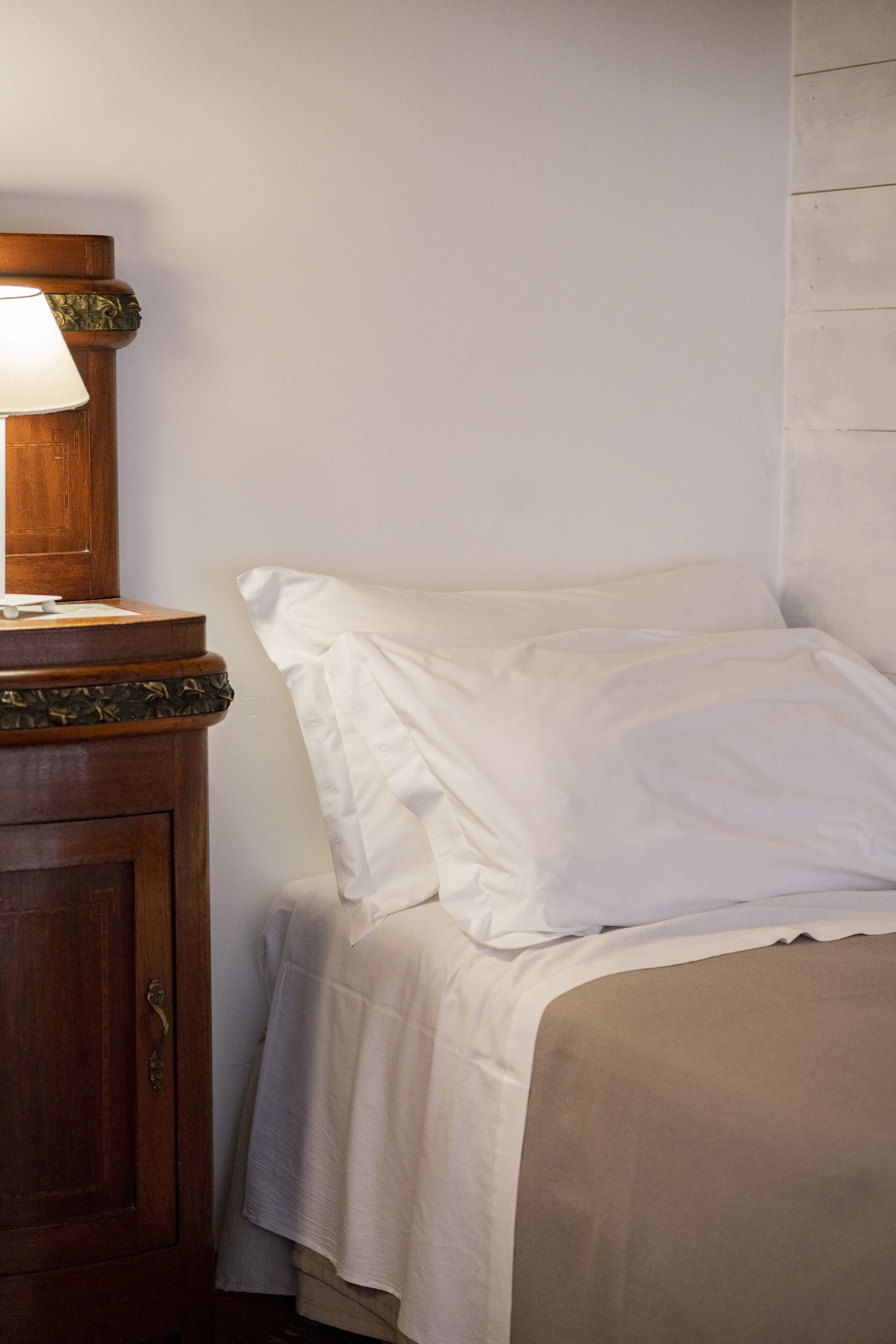 Quarto com uma cama, uma mesa de cabeceira de madeira com uma luminária e uma parede lisa.