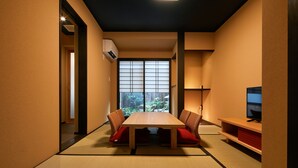 Um quarto tradicional japonês com piso de tatame, uma mesa baixa com almofadas, uma televisão em uma prateleira e uma janela com vista para o verde.