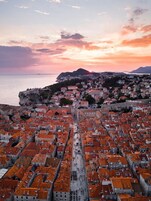 Uma vila costeira com edifícios de telhados vermelhos, uma rua central e montanhas ao fundo durante o pôr do sol.
