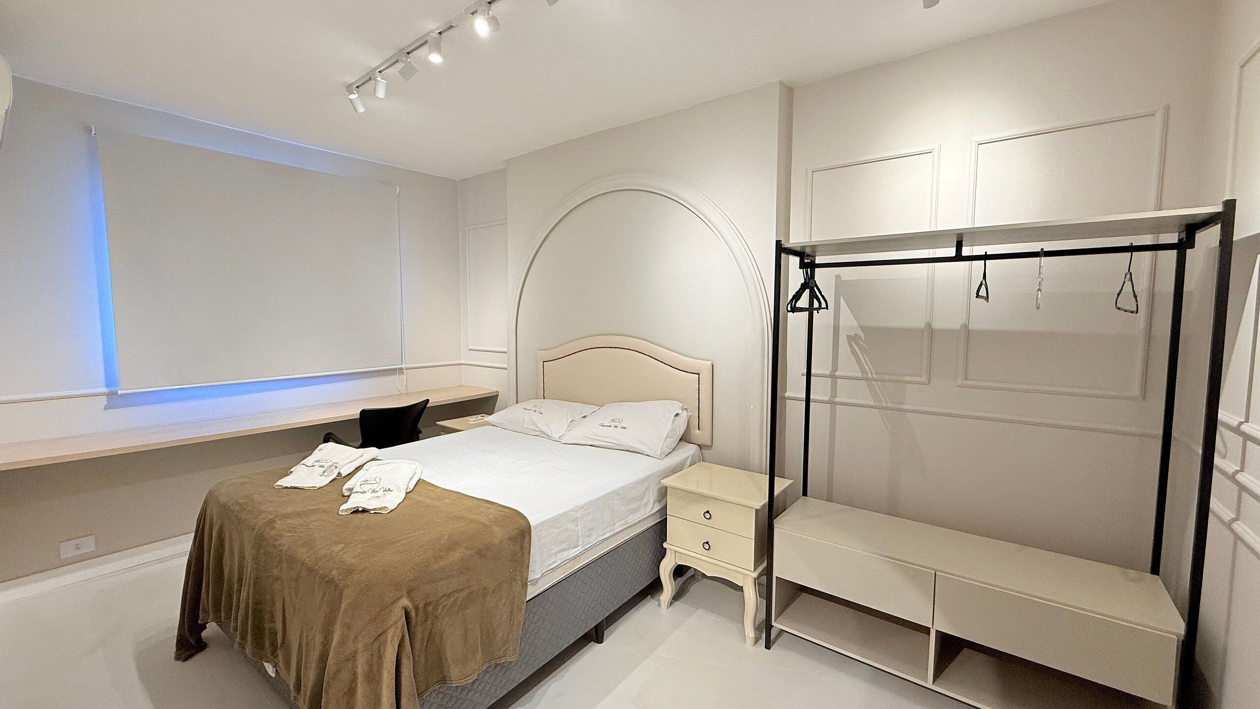 Quarto com cama, mesas de cabeceira, um banquinho e um guarda-roupa com espelho.