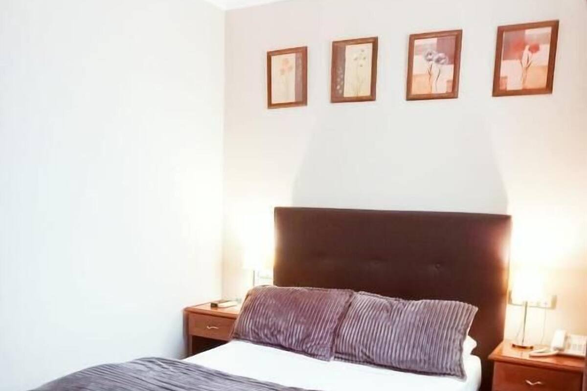 Quarto com cama, mesa de cabeceira e luminária na parede.