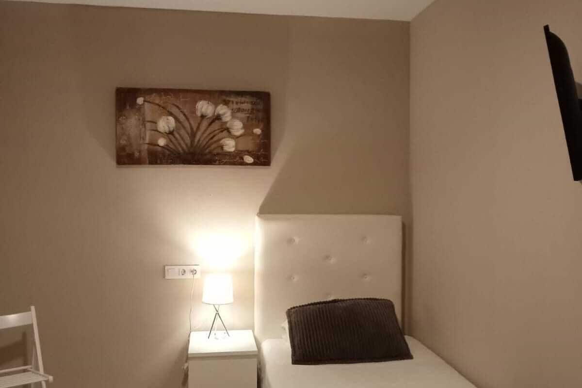 Quarto pequeno e bem arrumado, com uma cama de casal, mesa de cabeceira com luminária e uma obra de arte na parede.