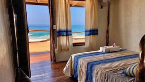 Quarto com cama, piso de madeira e vista para a praia através das cortinas.
