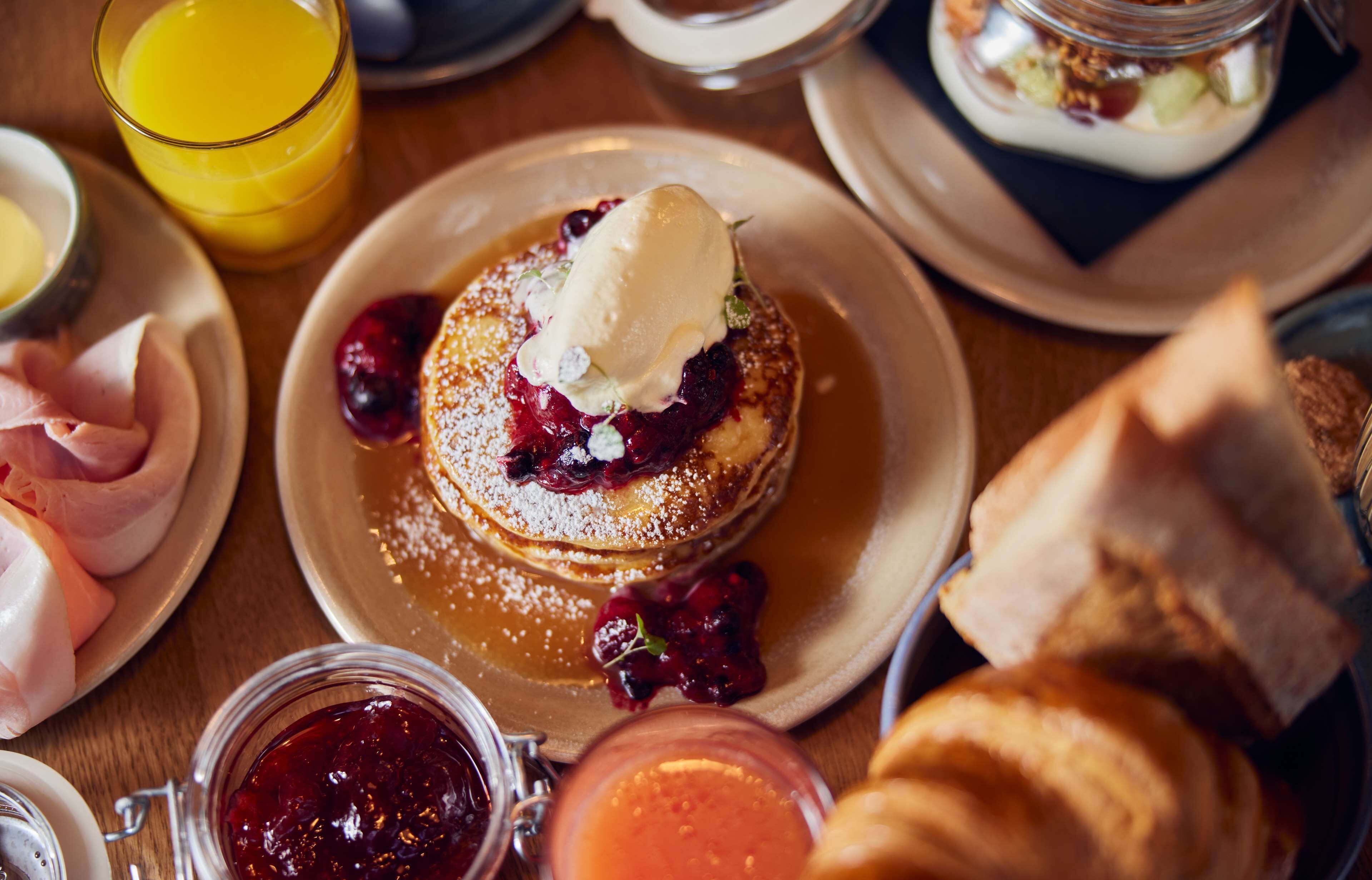 Uma mesa de café da manhã com panquecas cobertas de sorvete e frutas vermelhas, acompanhadas de torradas e um copo de suco de laranja.