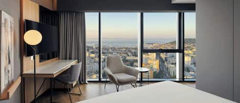 Quarto de hotel moderno com uma cama grande, uma escrivaninha com cadeira, televisão e vista para o horizonte urbano através de amplas janelas.