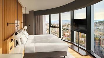 Quarto moderno com uma cama grande, televisão e vista para a cidade através de amplas janelas.
