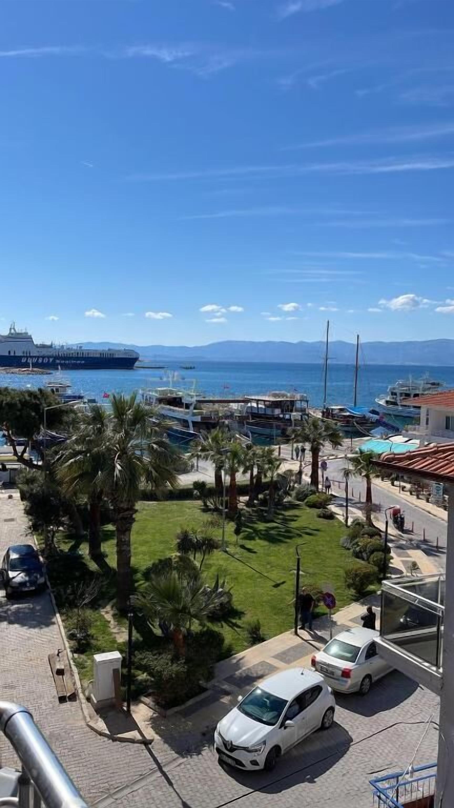 Vista costeira com um grande navio atracado no porto, uma marina com barcos e uma área de estacionamento com carros.