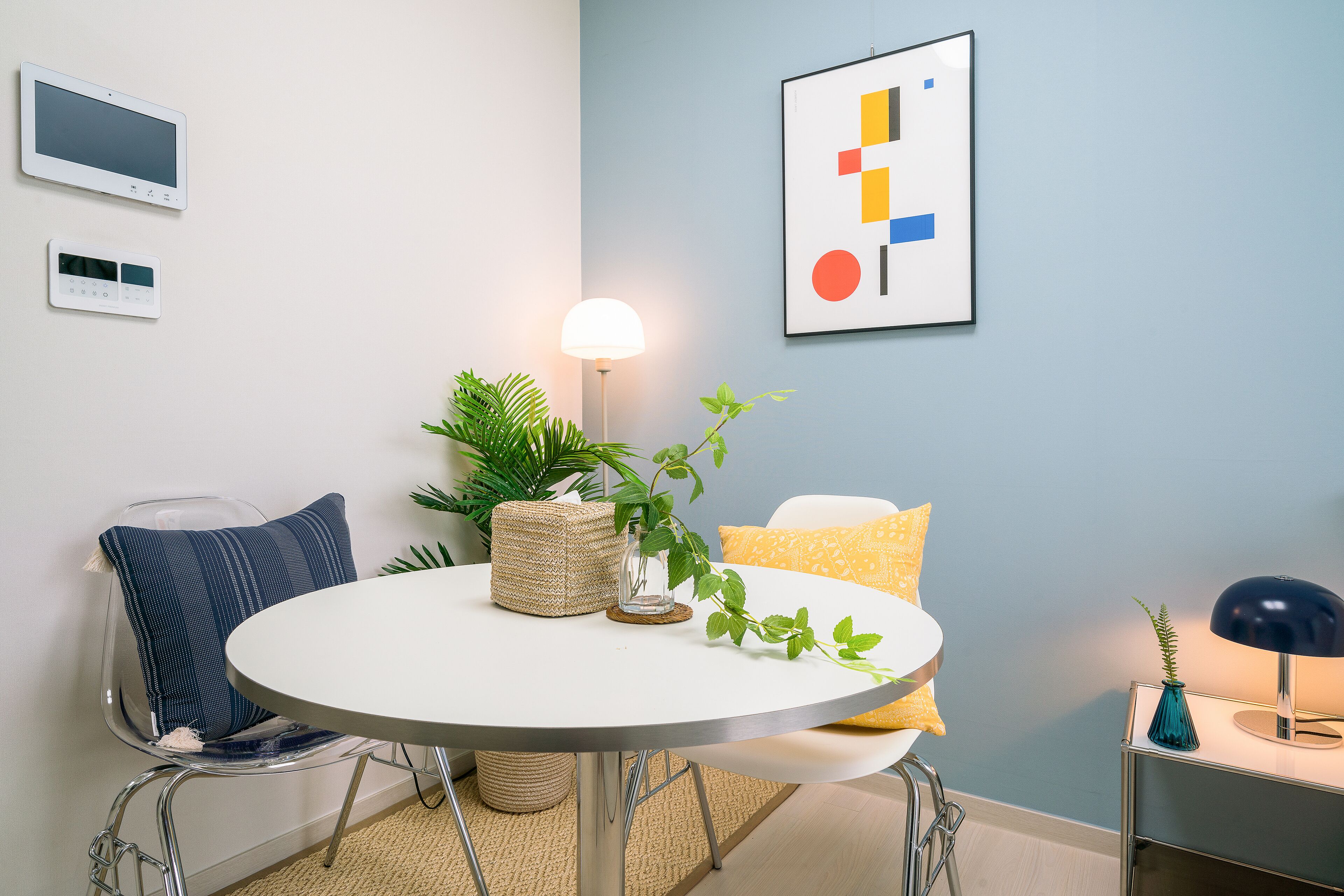 Quarto moderno com mesa redonda, cadeira transparente e abajur. Há uma planta na mesa e um painel de controle fixado na parede.