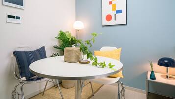Quarto moderno com mesa redonda, cadeira transparente e abajur. Há uma planta na mesa e um painel de controle fixado na parede.