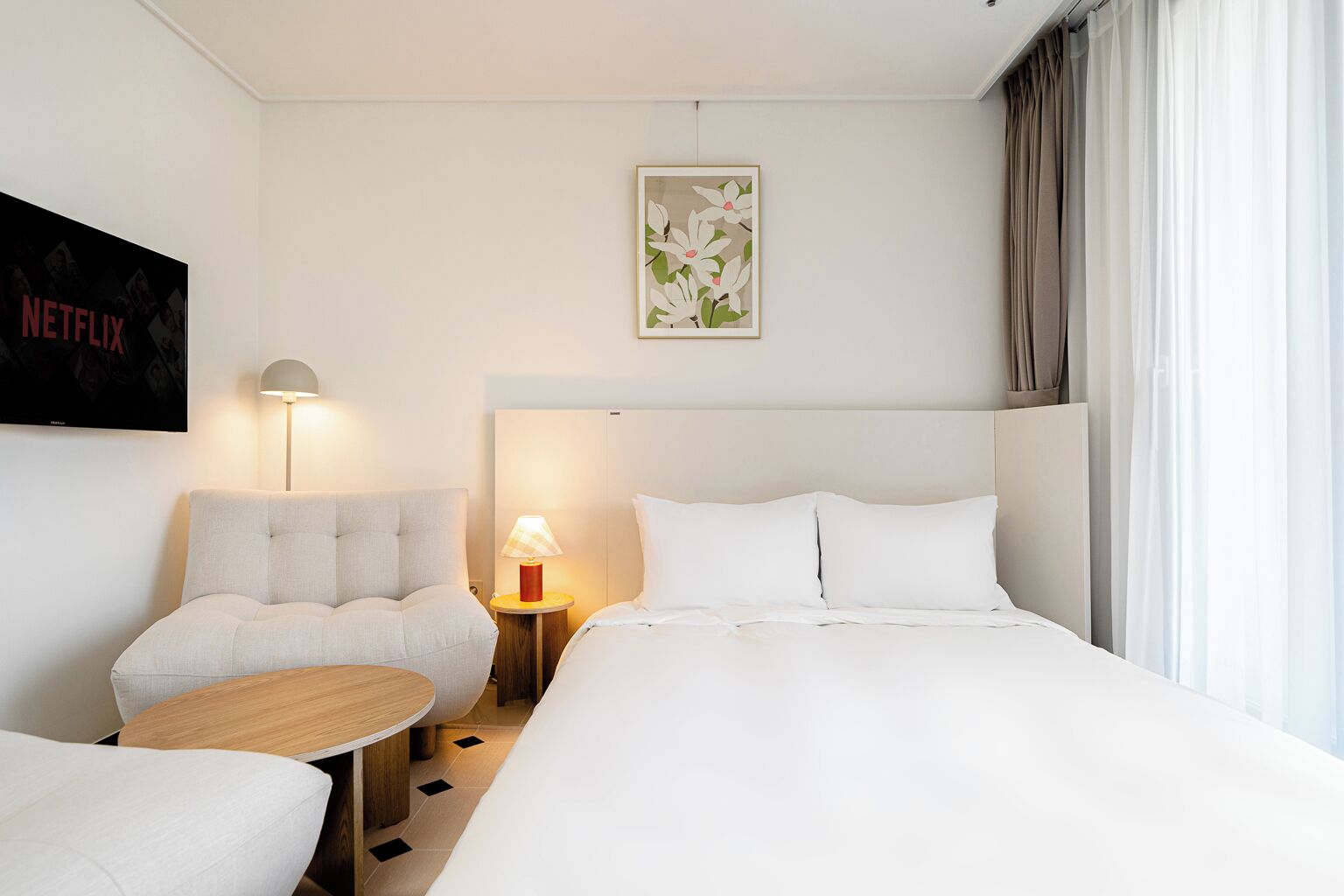 Quarto de hotel com cama, cadeira, mesa pequena, luminária e quadro pendurado na parede.