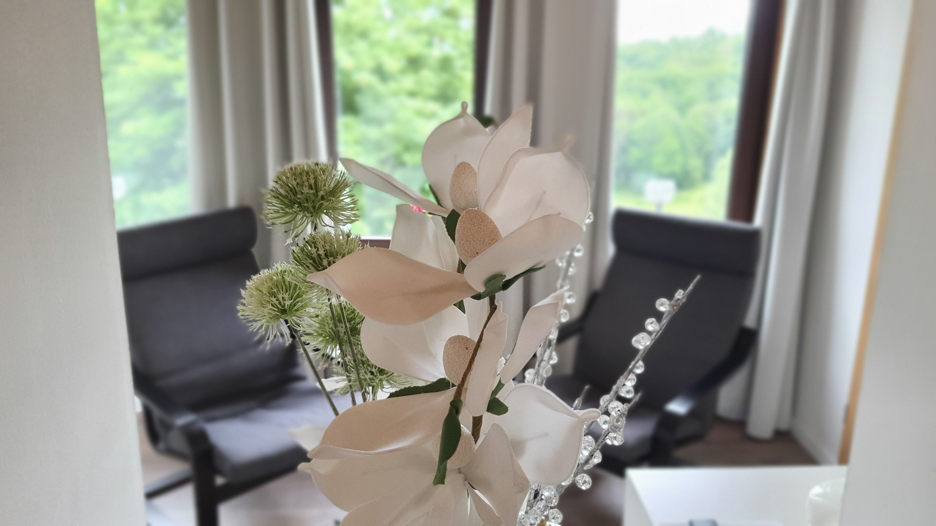 Um quarto com uma janela grande, um sofá preto e uma planta decorativa com flores brancas e folhagem verde.