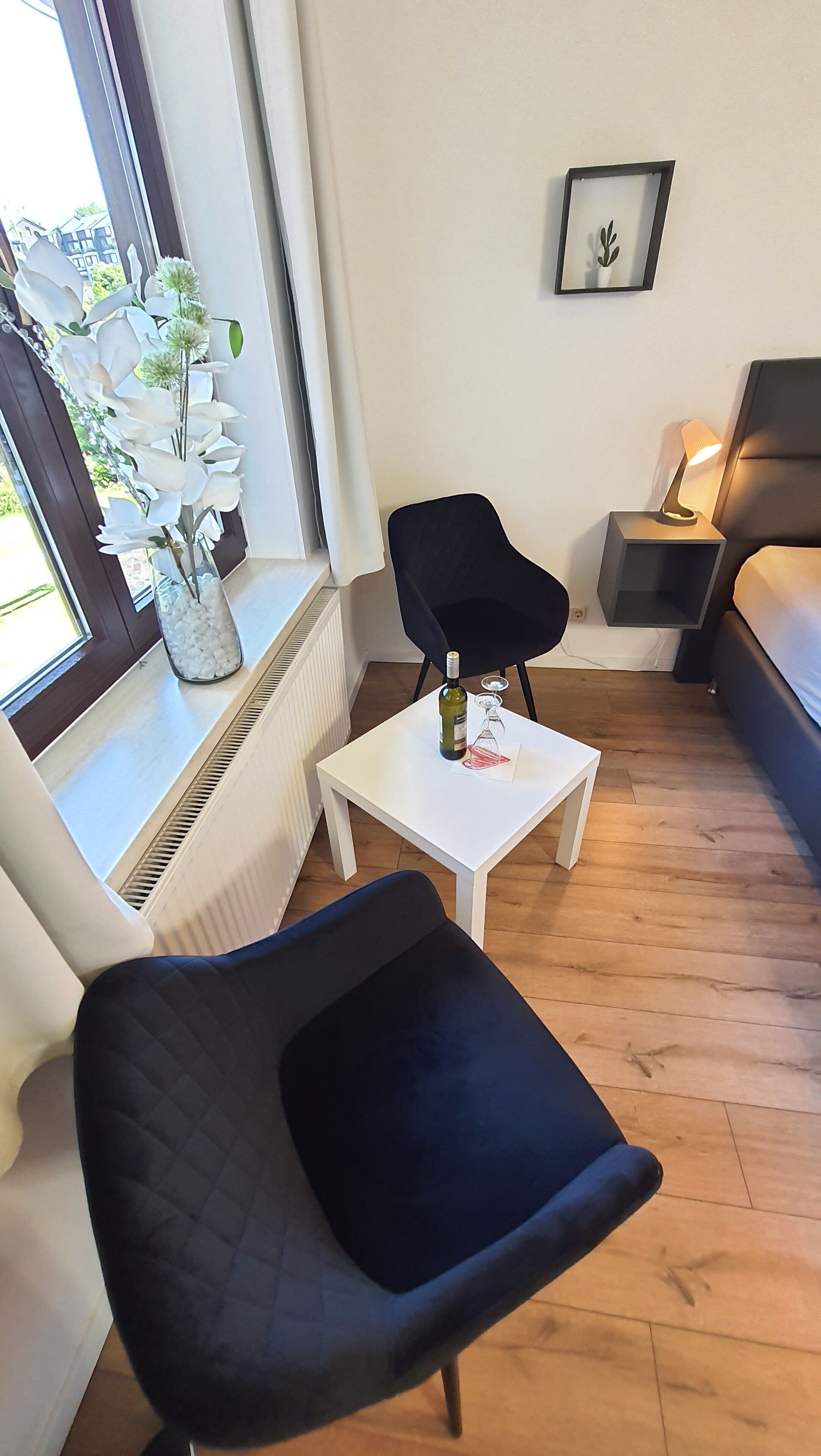 Uma sala de estar moderna com uma cadeira preta, uma mesa branca e uma pequena mesa lateral com uma luminária. Há uma garrafa de vinho e um copo sobre a mesa.